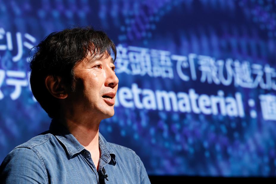 A man standing facing right in a blue shirt in front of a visual display during a presentation in front of an audience
